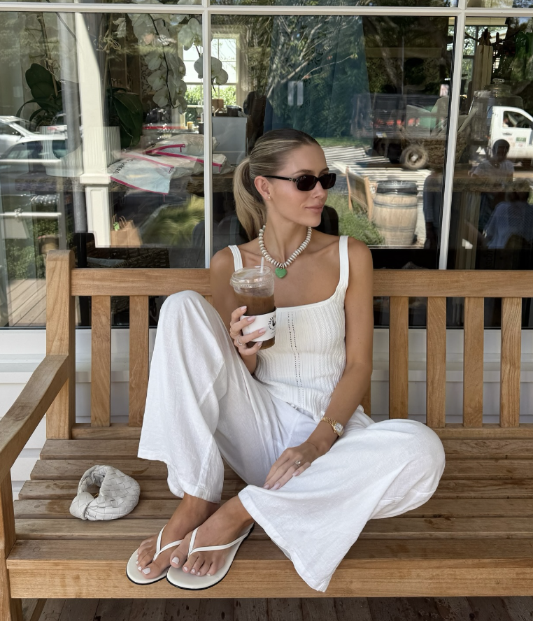 A woman in a white outfit and sunglasses sits on a wooden bench%2C holding an iced coffee. The background features large windows reflecting trees.